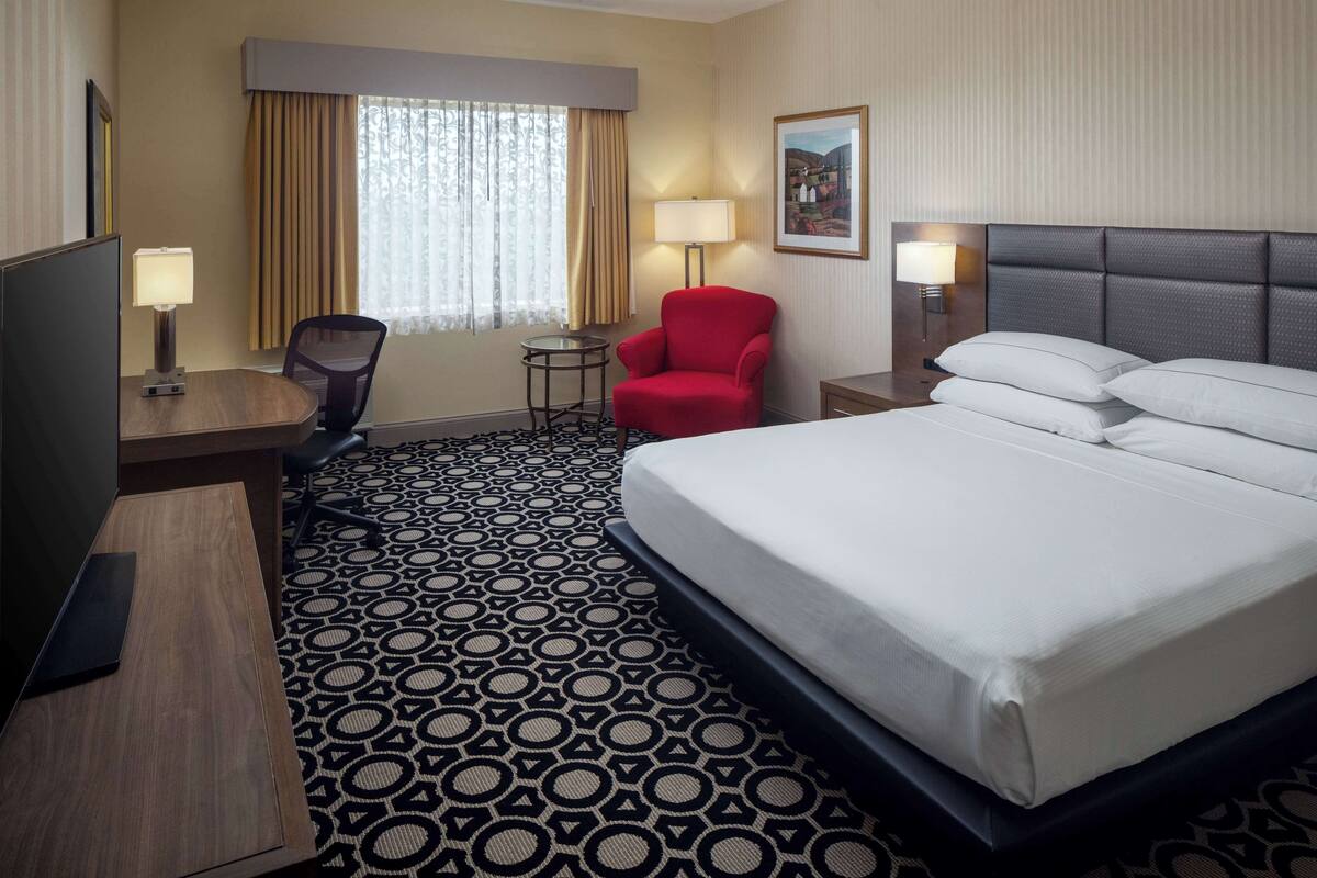 Quarto de hotel com uma cama grande, uma mesa com luminária, uma cadeira vermelha, uma mesinha e uma janela com cortinas.