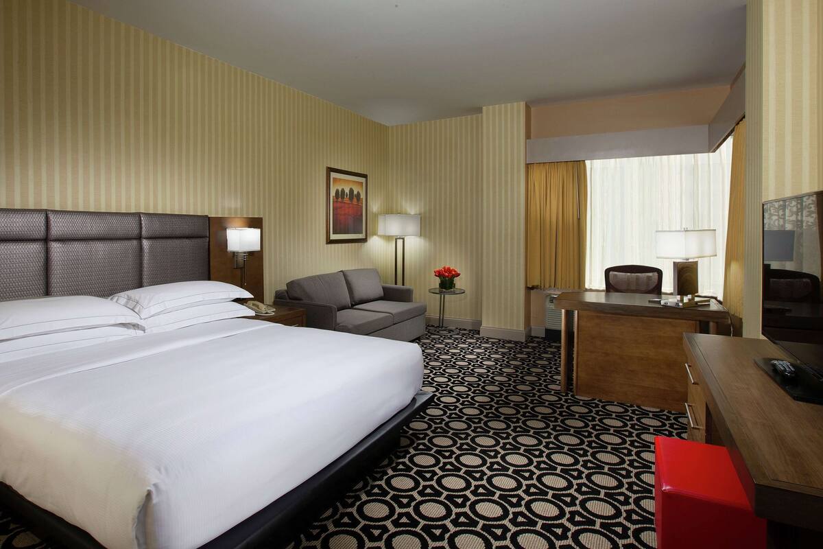 Quarto de hotel com uma cama grande, uma escrivaninha, uma cadeira, um sofá e uma televisão.