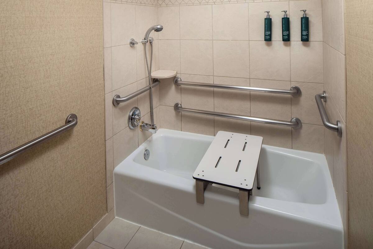 Casa de banho com banheira branca, barras de apoio, chuveiro manual e três frascos de produtos de higiene pessoal na parede.