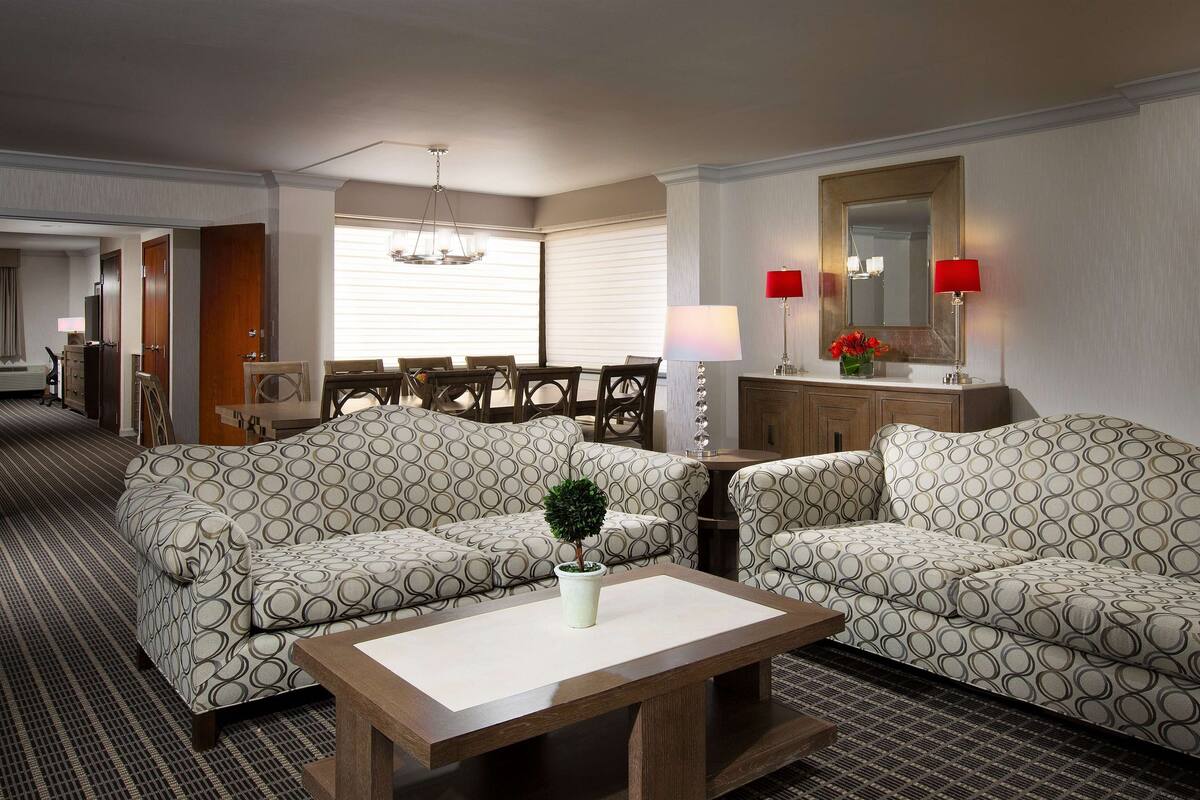 Quarto de hotel com sofá estampado, mesa de centro de madeira, área de refeições com cadeiras e espelho sobre a pia.