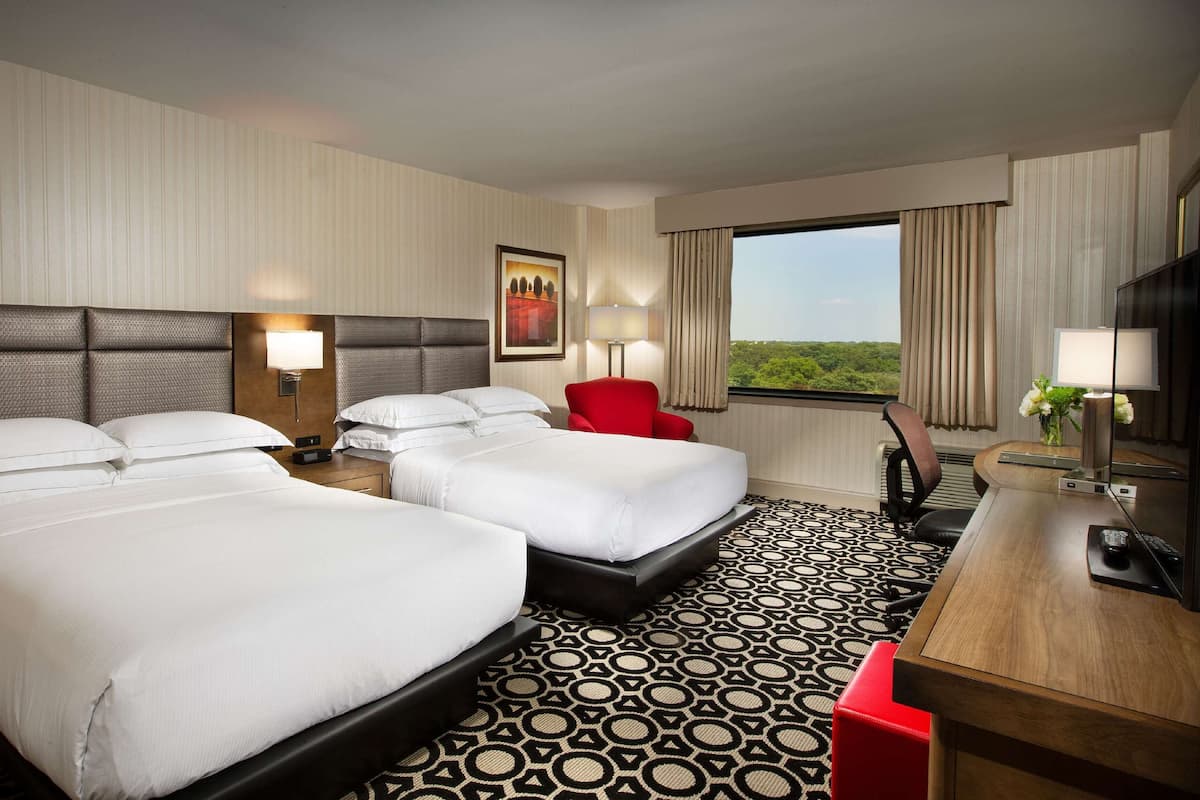 Quarto de hotel com duas camas, uma mesa, uma cadeira e uma janela com vista.