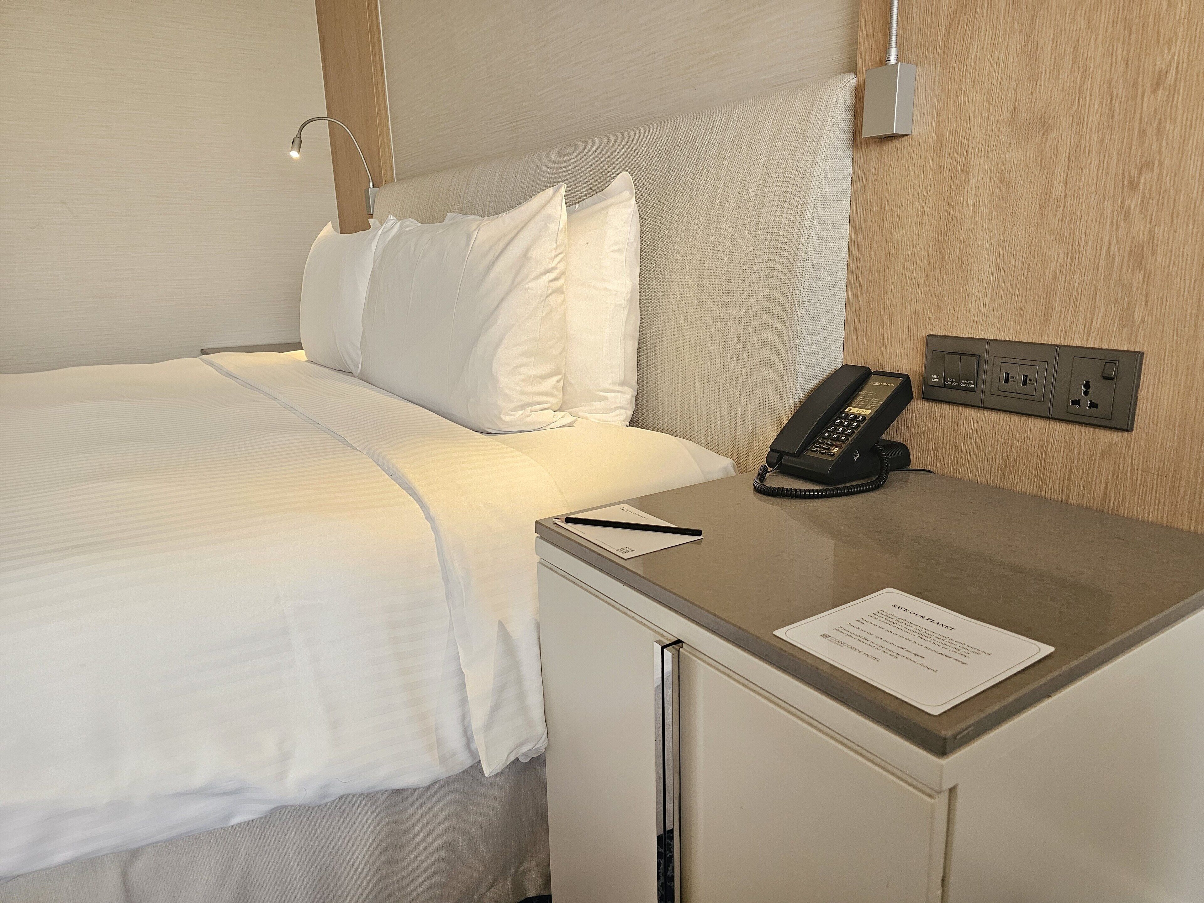 Quarto de hotel com cama bem arrumada, mesa de cabeceira com telefone e bloco de notas, e tomada na parede.
