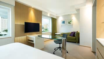 Quarto de hotel moderno com uma cama grande, uma escrivaninha de vidro com cadeira, televisão de tela plana, sofá verde e vista para a cidade.