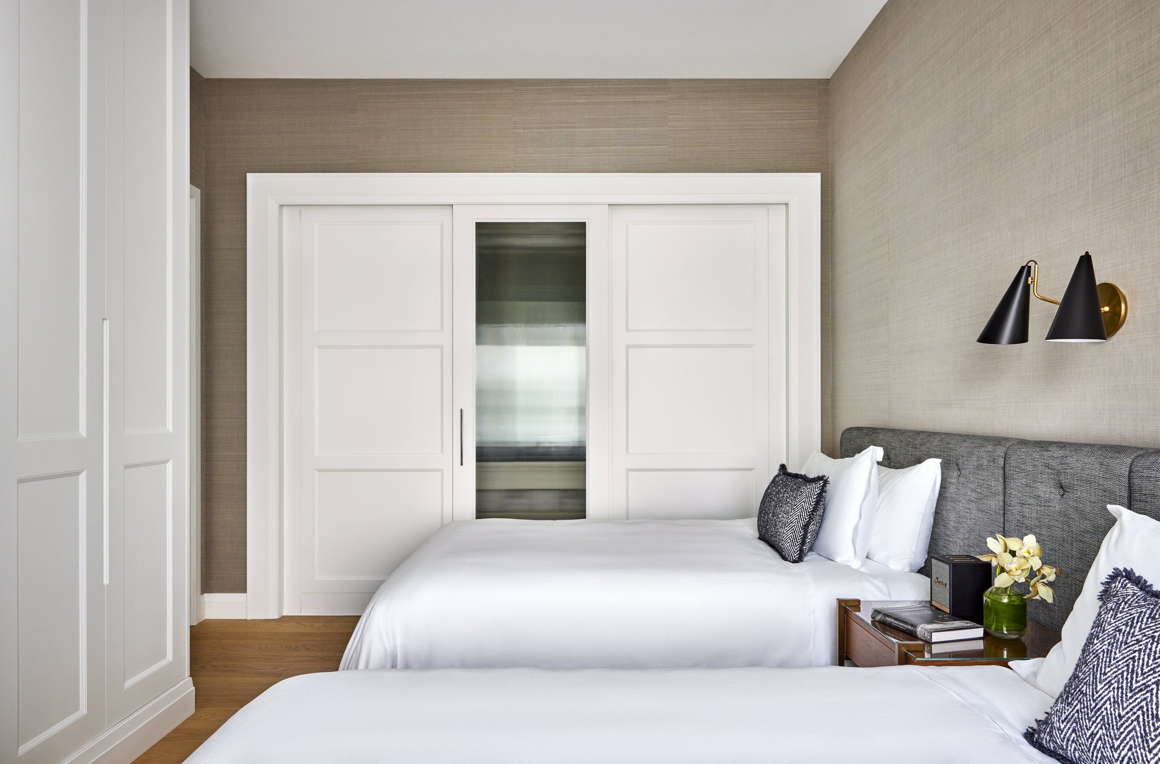 Quarto com duas camas, guarda-roupa, mesa de cabeceira com abajur e vista através de uma porta.