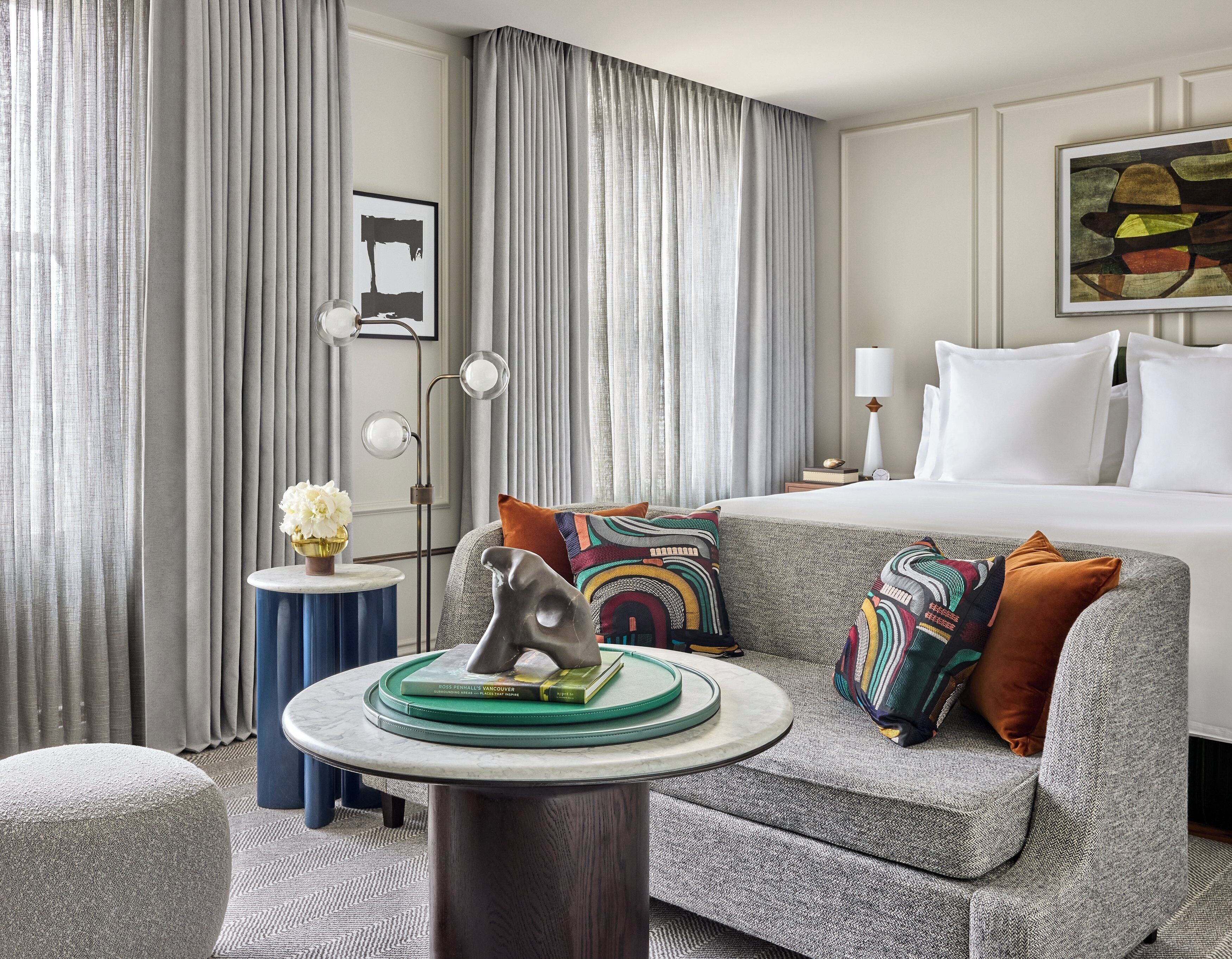 Quarto de hotel com cama, sofá, mesa redonda com uma escultura, luminária e um quadro na parede.