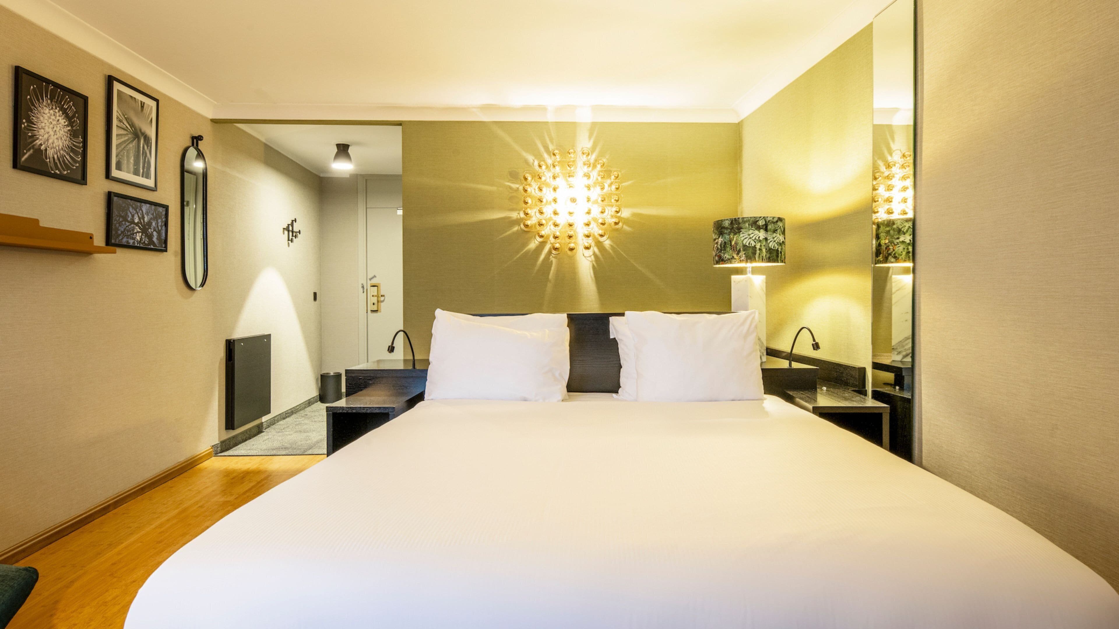 Quarto de hotel com uma cama grande, mesas de cabeceira, um espelho e quadros nas paredes.