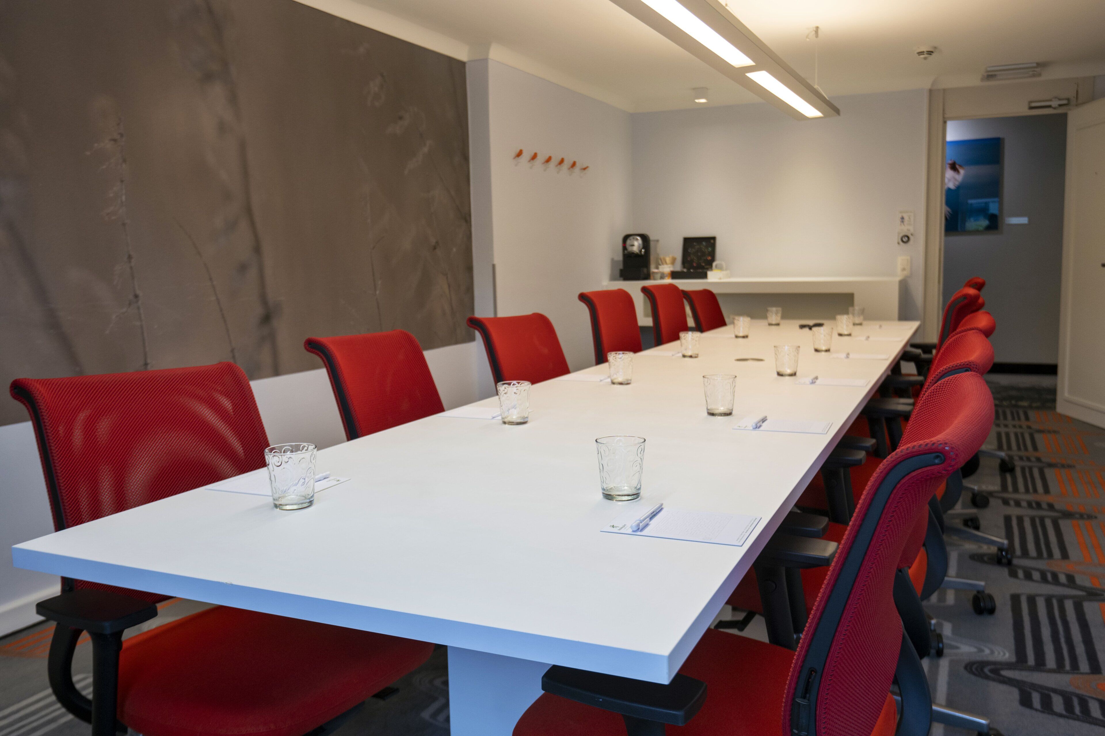 Uma sala de conferências com uma mesa comprida, cadeiras vermelhas e um quadro branco na parede.