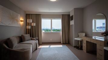 Quarto de hotel moderno com sofá, escrivaninha, espelho e vista panorâmica da cidade através de grandes janelas.