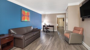 Quarto de hotel com sofá de couro marrom, escrivaninha com luminária, TV fixada na parede e piso de madeira.