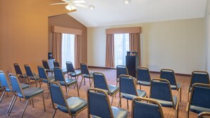 Uma sala de conferências com fileiras de cadeiras azuis, ventiladores no teto e amplas janelas com cortinas.