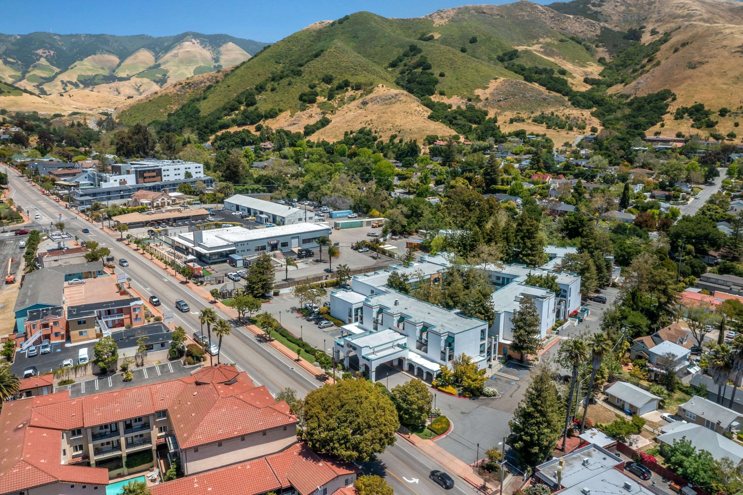 Vista aérea de uma cidade com mistura de edifícios modernos e tradicionais, estradas e colinas verdes ao fundo.
