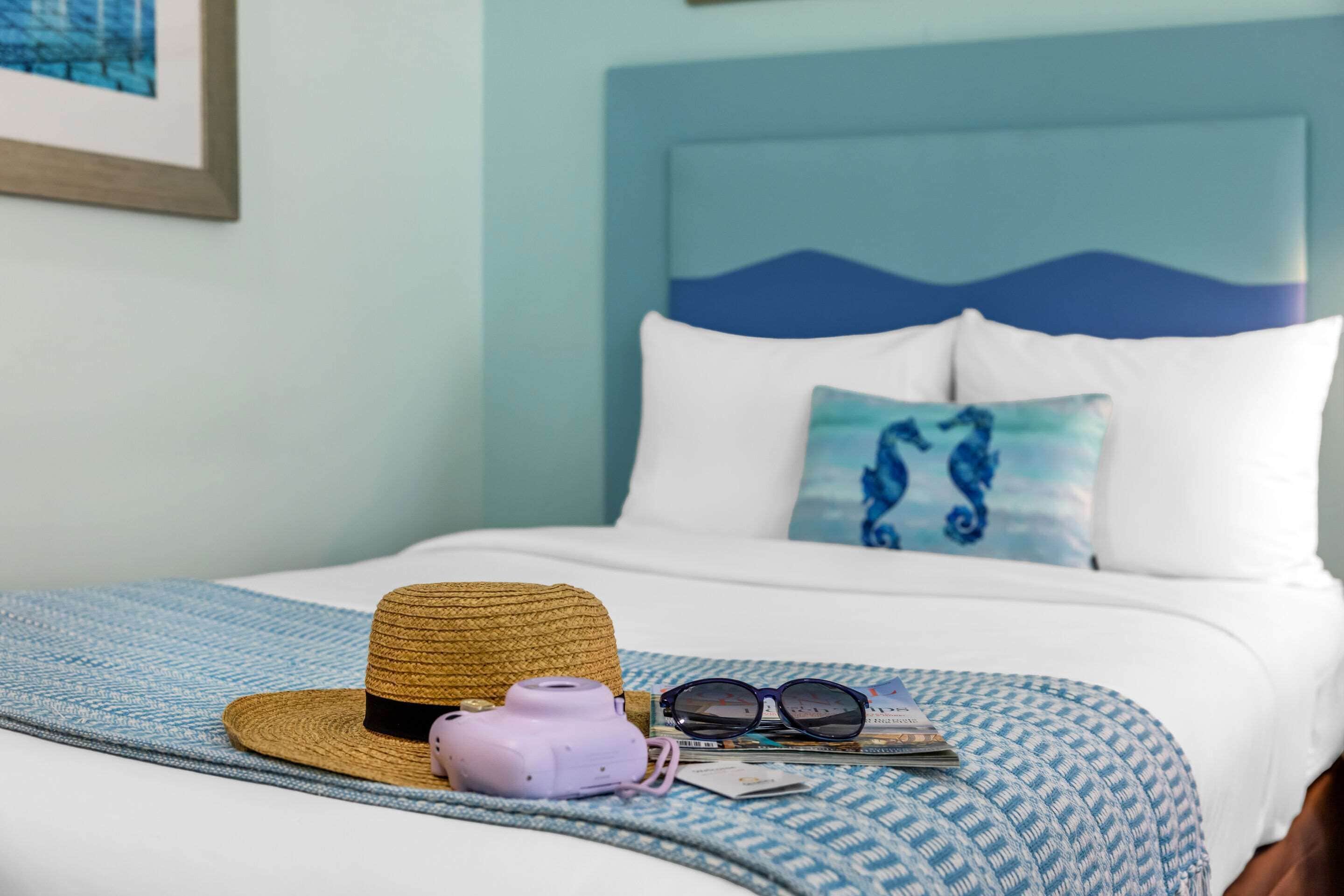 Uma cama bem arrumada com um cobre-leito estampado em azul e branco, um chapéu de palha, um par de óculos de sol e uma câmera sobre a cama.