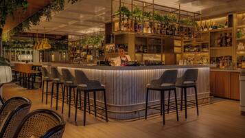 Uma área de bar com balcão curvo, banquetas, prateleiras com garrafas e plantas penduradas.