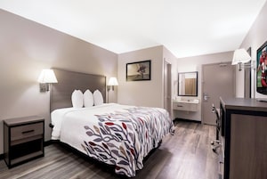 Quarto de hotel com cama, luminárias de cabeceira, um quadro, banheiro com pia e espelho, e televisão.