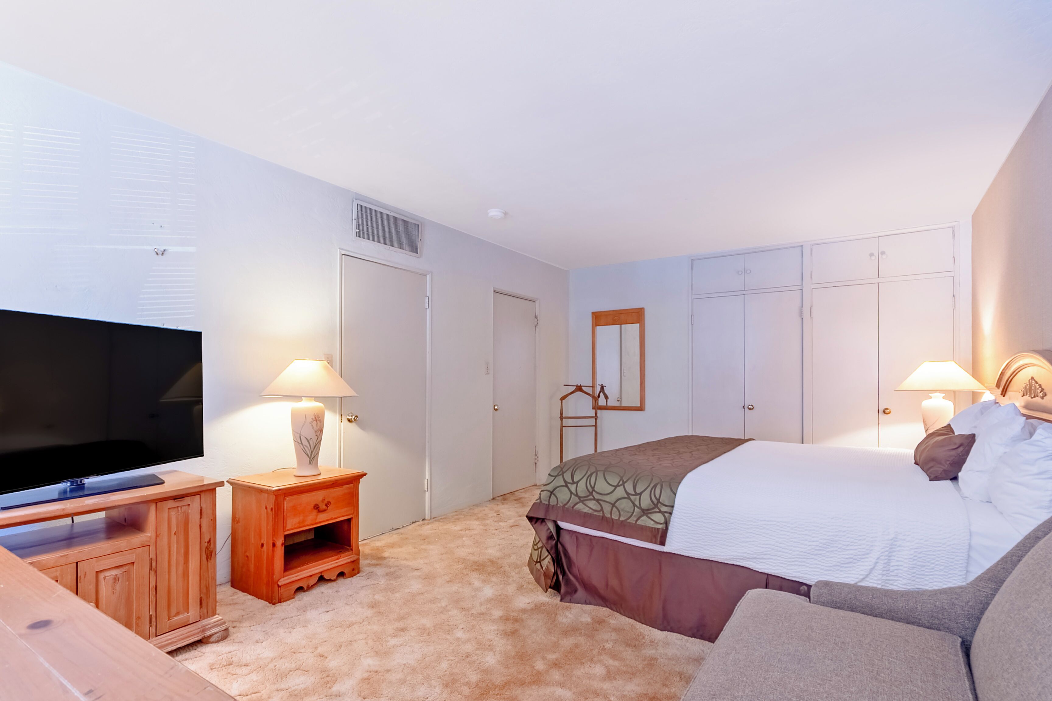 Quarto de hotel com uma televisão de tela plana grande, mesas de cabeceira de madeira, uma cama com colcha estampada e um guarda-roupa com várias portas.