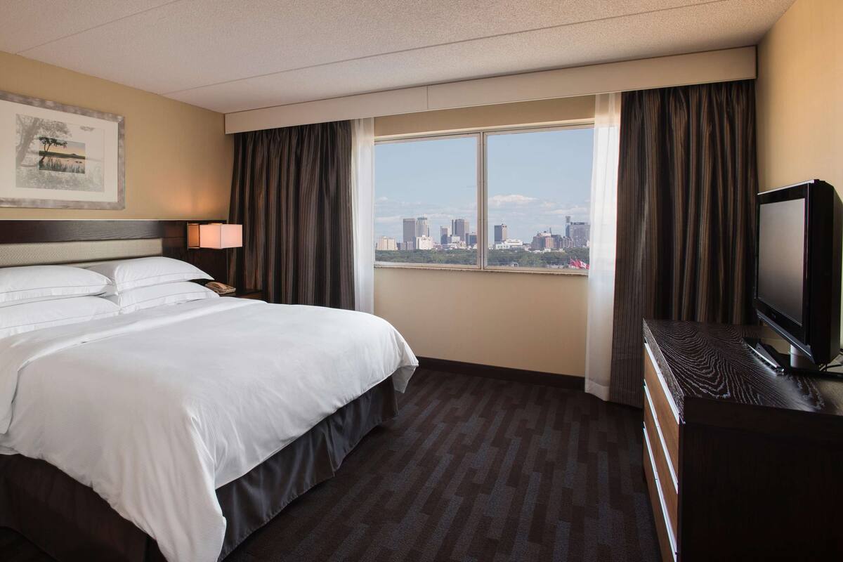 Quarto de hotel com uma cama grande, uma escrivaninha com luminária, uma televisão e uma janela com vista para a cidade.