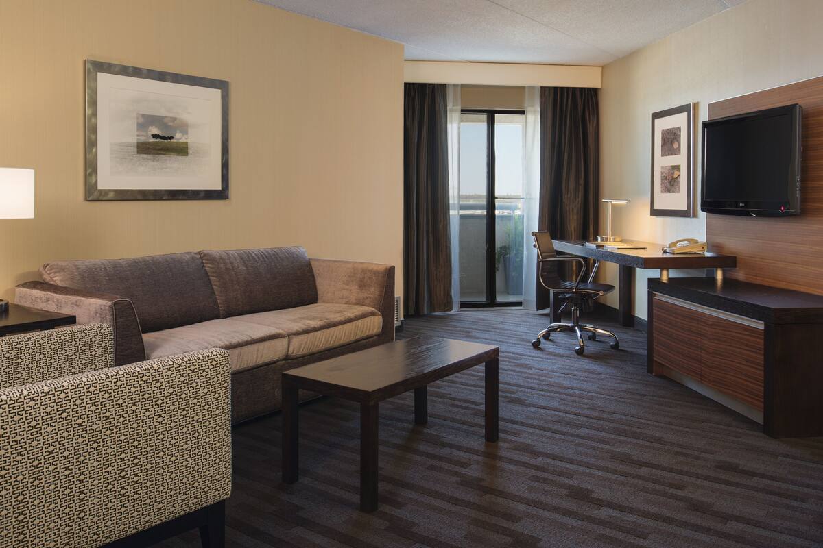 Quarto de hotel com sofá, poltrona, escrivaninha e televisão.