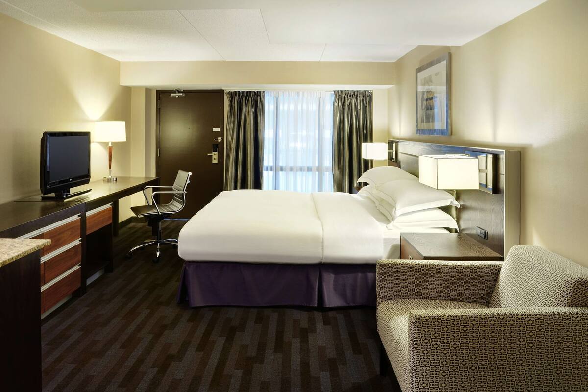 Quarto de hotel com cama, escrivaninha, cadeira, televisão e luminárias.