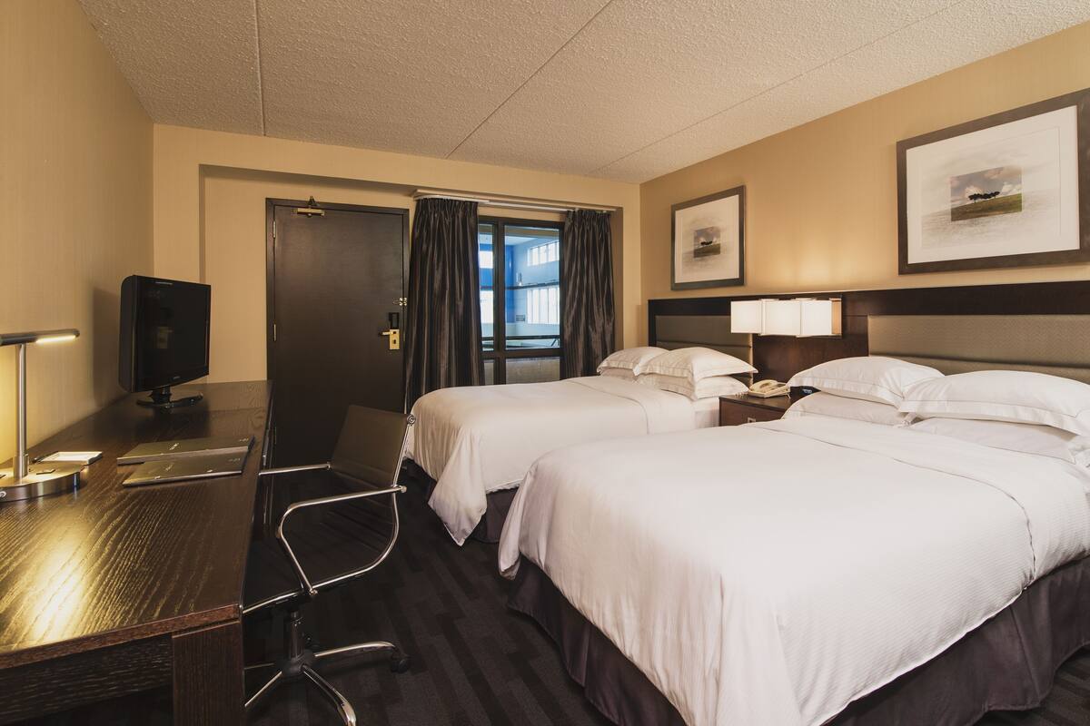 Quarto de hotel com duas camas, uma escrivaninha com computador, televisão e janela com cortinas.