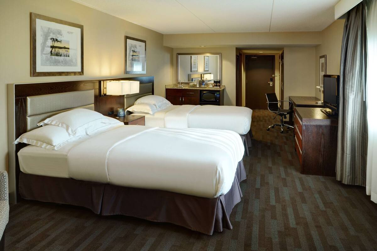 Quarto de hotel com duas camas grandes, uma escrivaninha, uma cadeira, televisão e banheiro.