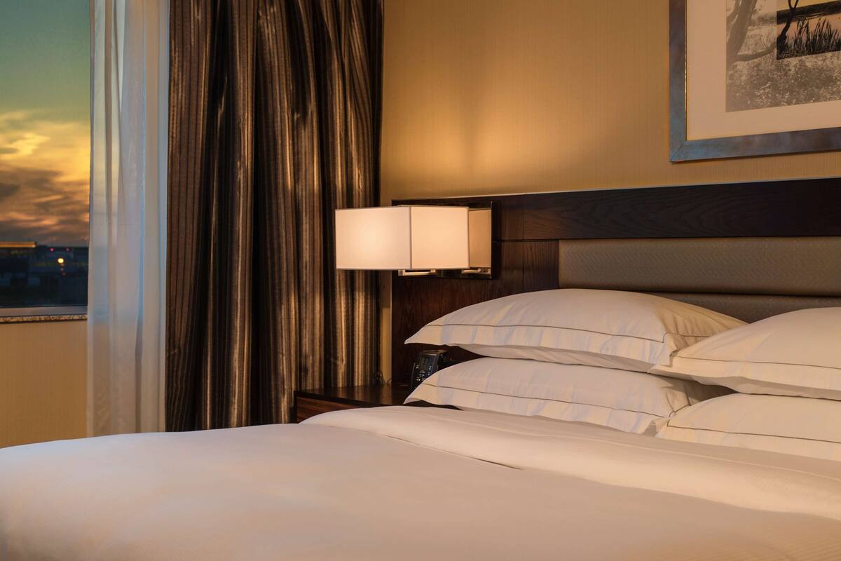 Quarto de hotel com uma cama grande, luminária de cabeceira e vista para a cidade ao entardecer.