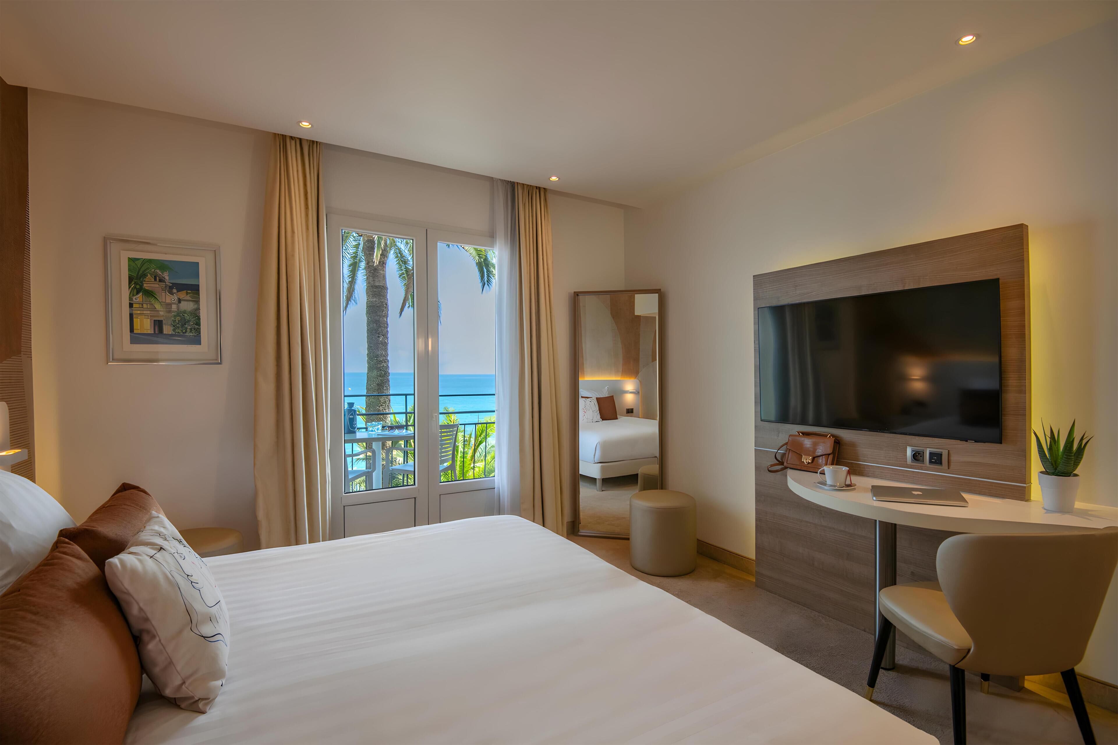 Quarto de hotel com uma cama grande, uma escrivaninha com um laptop, uma TV e uma varanda com vista para o mar.