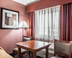 Quarto de hotel com mesa de madeira, duas cadeiras, abajur, janela grande com cortinas translúcidas e quadro decorativo na parede.