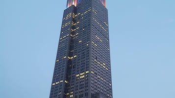 Um arranha-céu alto e moderno, com um pináculo característico e janelas iluminadas, rodeado por outros edifícios altos.