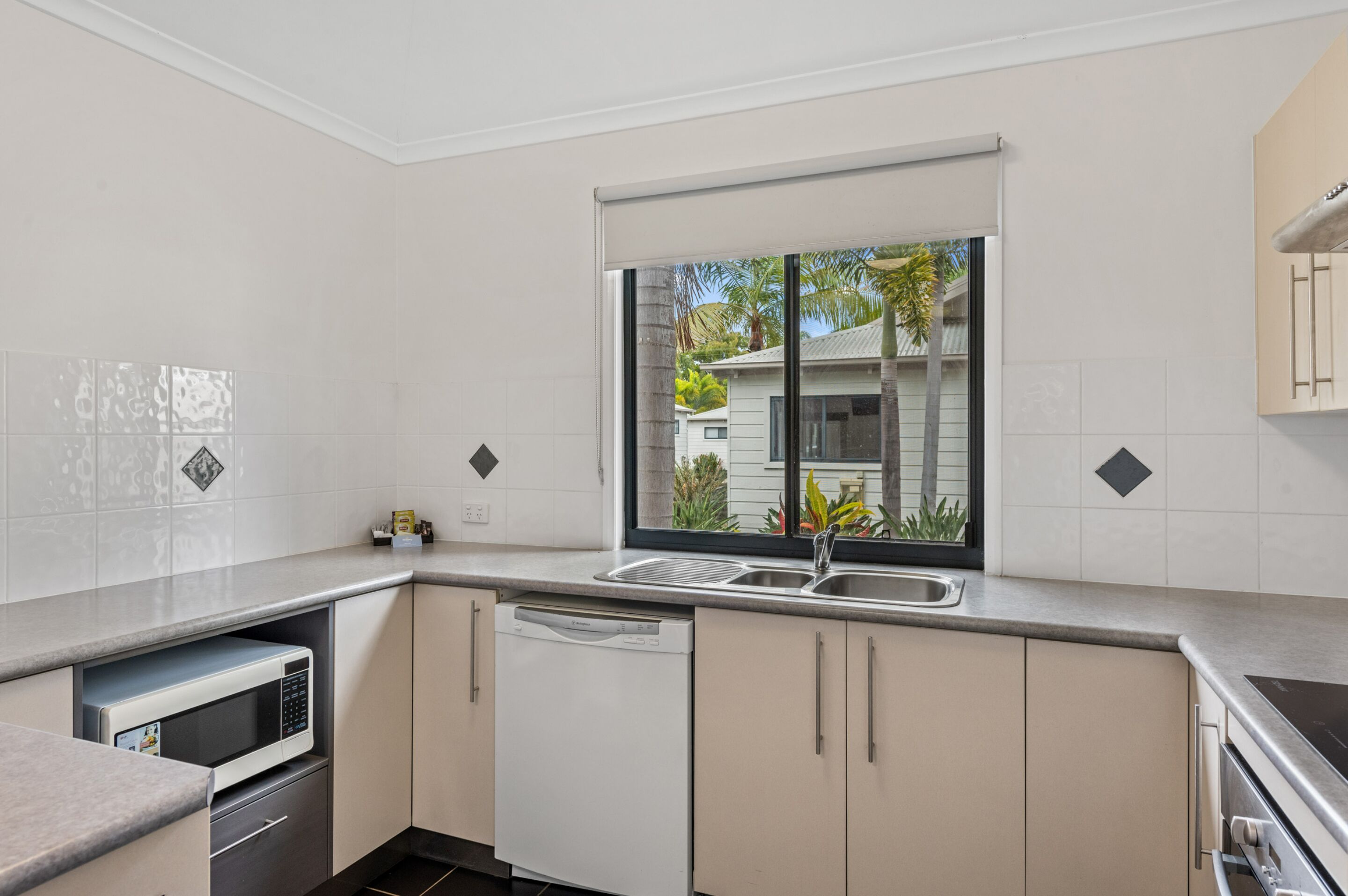 Uma cozinha moderna com armários brancos, lava-louças, micro-ondas e uma janela com vista para uma casa e palmeiras.