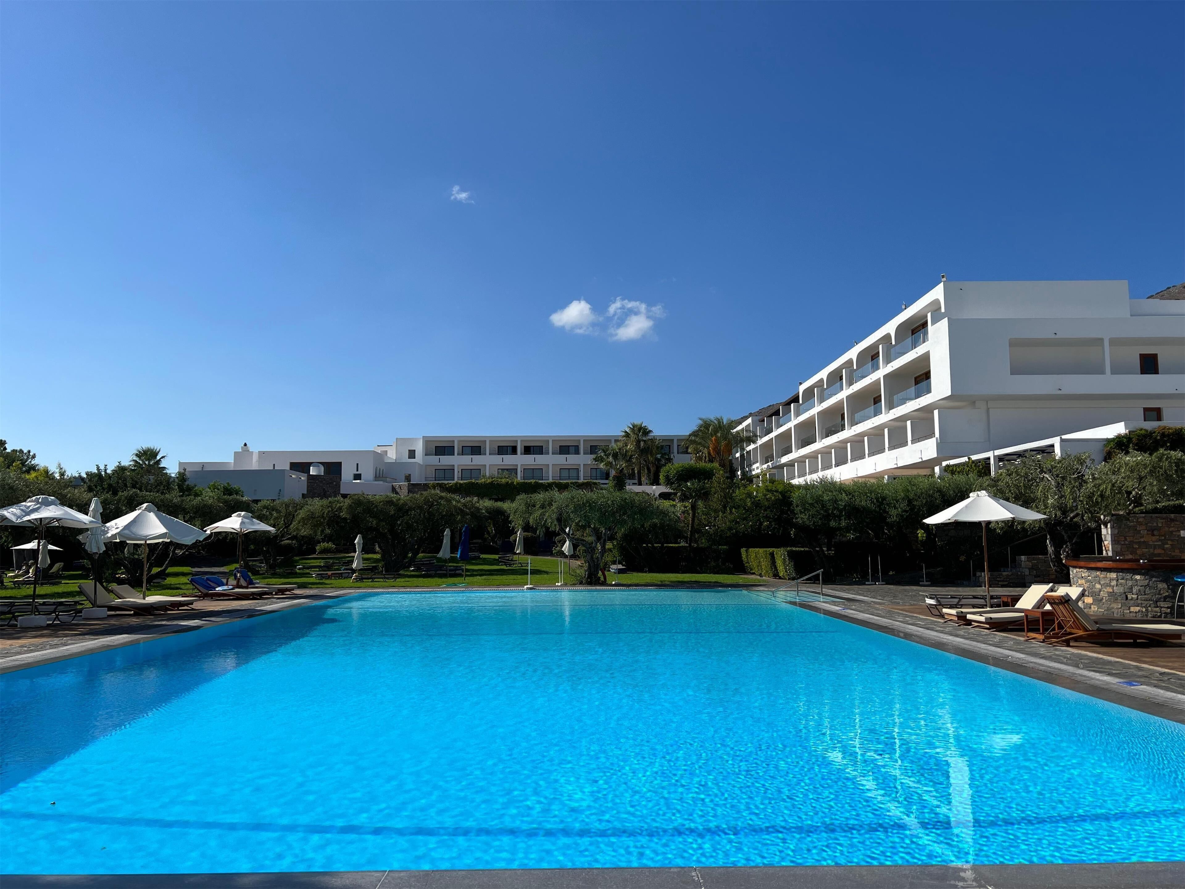 Piscina com espreguiçadeiras e guarda-sóis, cercada por vegetação e um edifício branco com várias varandas.