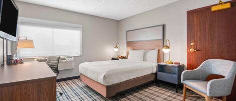 Quarto de hotel com uma cama, uma escrivaninha, uma cadeira, uma televisão, uma janela e uma porta.