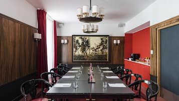 Uma sala de conferências com uma mesa comprida, cadeiras, um candelabro, um quadro e uma parede com detalhes em vermelho.