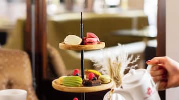 Suporte para sobremesas em três níveis com diversos doces e frutas, uma xícara branca e um pires sobre uma mesa.