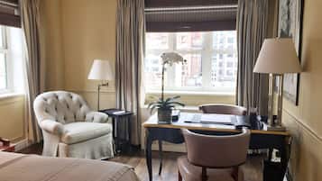 Quarto de hotel com cama, escrivaninha, cadeira, luminária, planta em vaso e janela com cortinas.