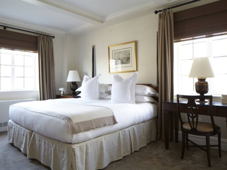 Quarto de hotel bem organizado, com uma cama grande, uma escrivaninha e uma cadeira. Há um quadro na parede e uma janela com cortinas.