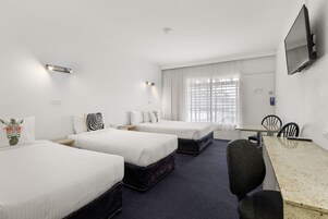 Quarto de hotel com três camas, uma escrivaninha, uma cadeira e televisão na parede.