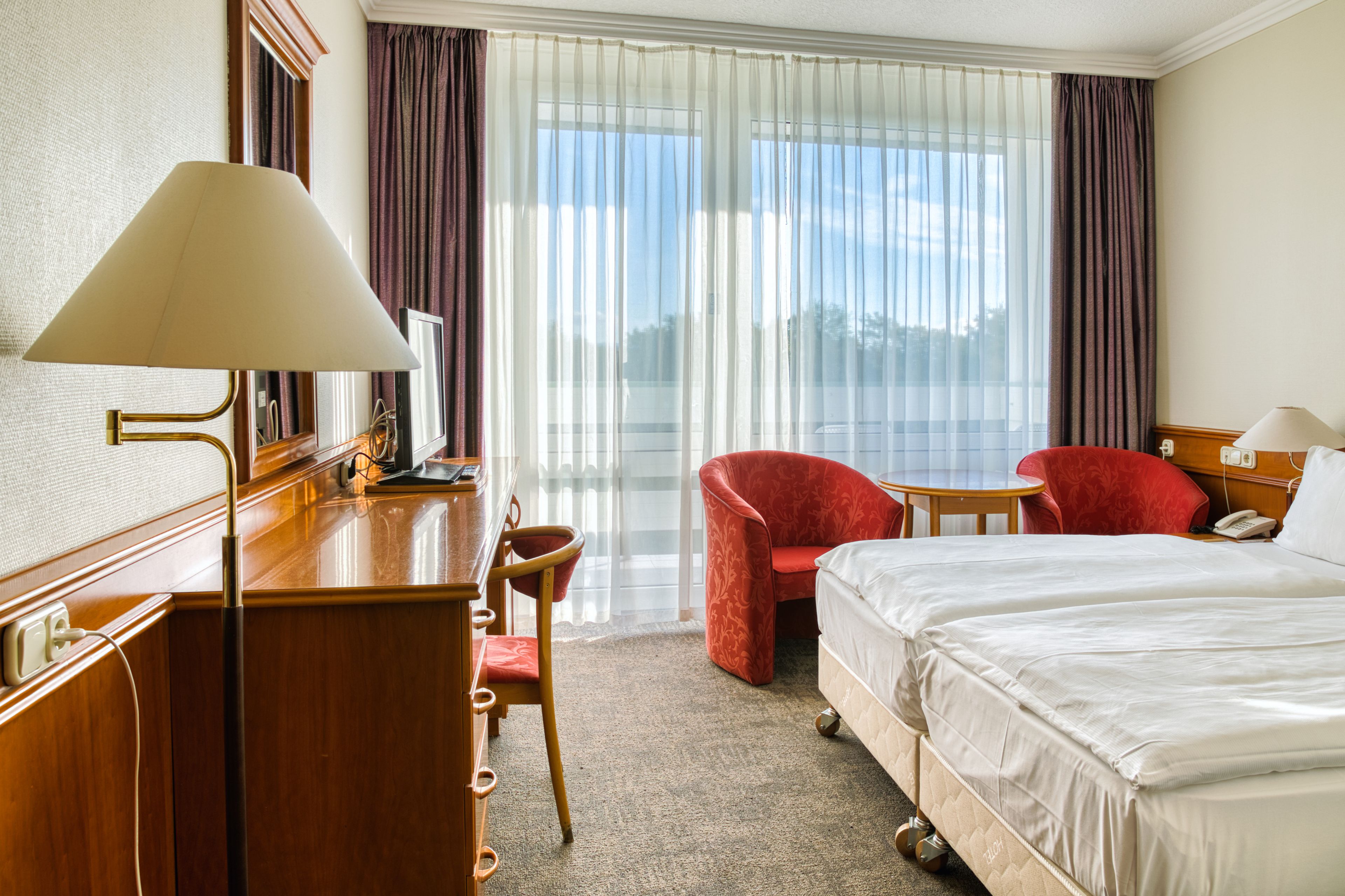 Quarto de hotel com cama, escrivaninha, cadeira, mesa e janela com cortinas.
