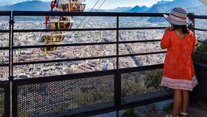 Um sistema de teleférico com passageiros, paisagem urbana ao fundo e uma pessoa perto de um corrimão.