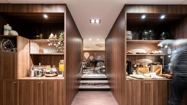 Corredor de hotel com painéis de madeira, escada central e área de café da manhã com comida e bebidas.