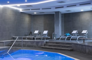Uma área interna de piscina moderna, com piscina cristalina, degraus para entrar na água e várias espreguiçadeiras com almofadas.