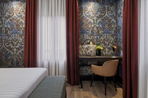 Quarto de hotel com cama, escrivaninha com cadeira, luminária e um vaso com flores.