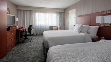 Quarto de hotel com duas camas, uma mesa de trabalho com televisão, uma cadeira e uma janela grande com cortinas.