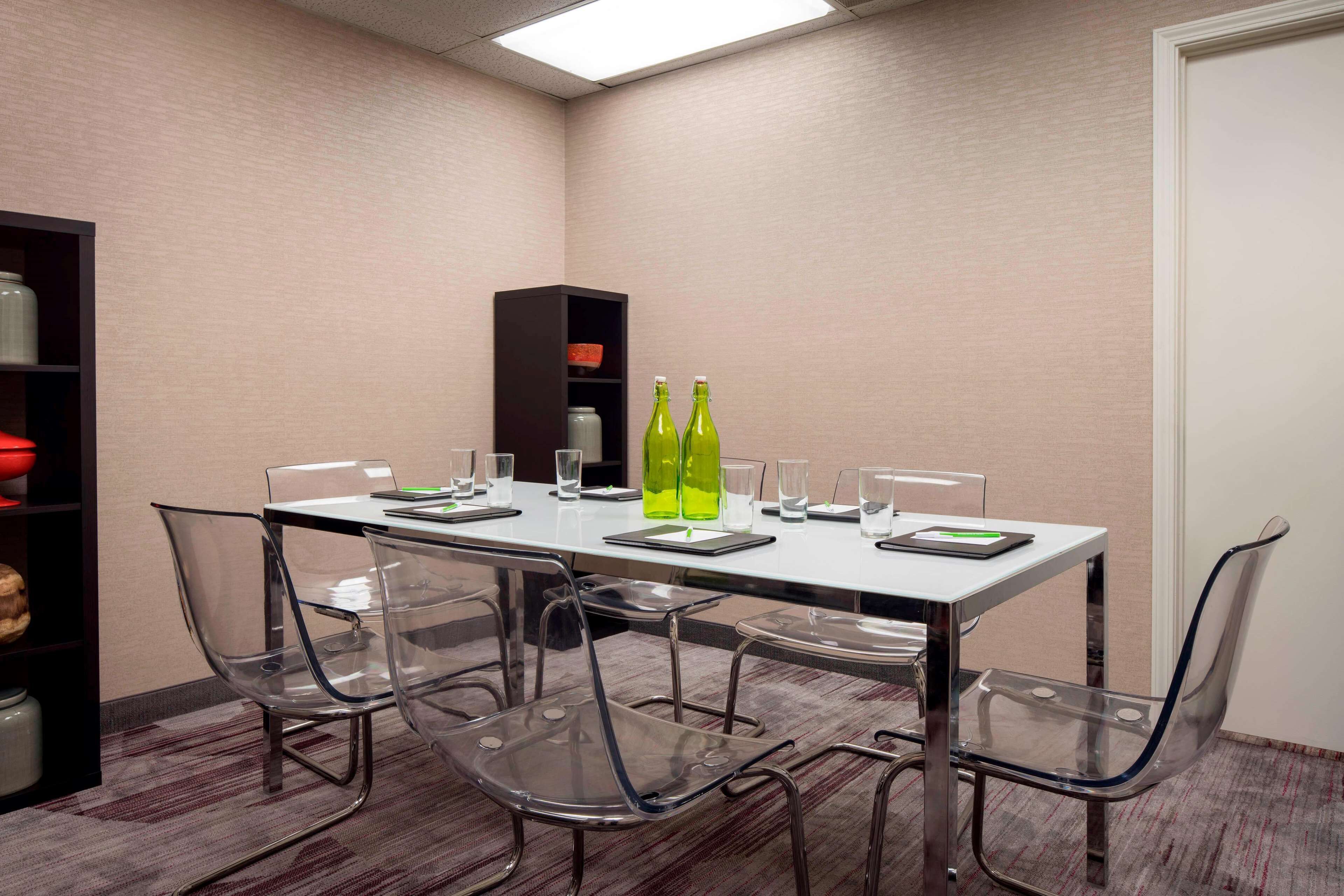 Uma sala de conferências com uma mesa branca, cadeiras transparentes e uma estante de livros.