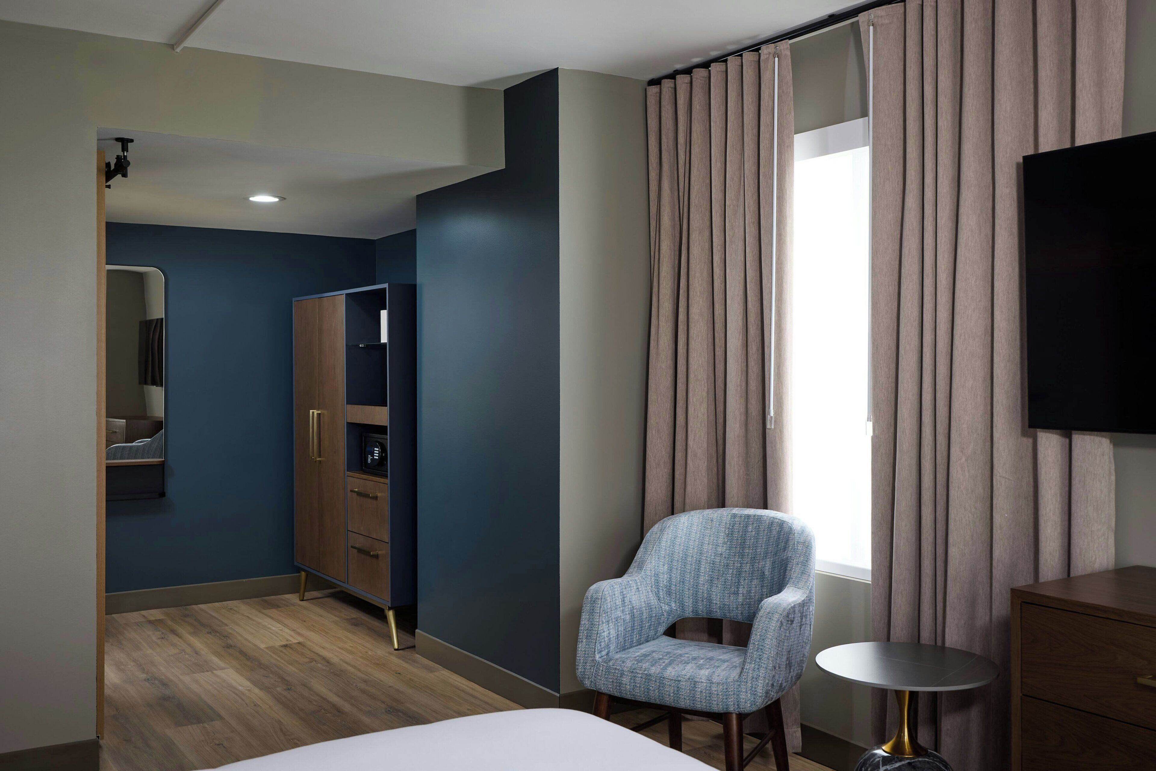 Quarto de hotel com cama, uma cadeira, uma mesinha, guarda-roupa e televisão na parede.