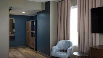 Quarto de hotel com cama, uma cadeira, uma mesinha, guarda-roupa e televisão na parede.