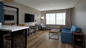 Quarto de hotel moderno com piso de madeira, TV de tela plana na parede, escrivaninha com cadeira, sofá e uma pequena mesa com abajur.