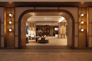 Um hall de hotel com design moderno, com área de estar central, balcão de recepção e uma grande tela de televisão.