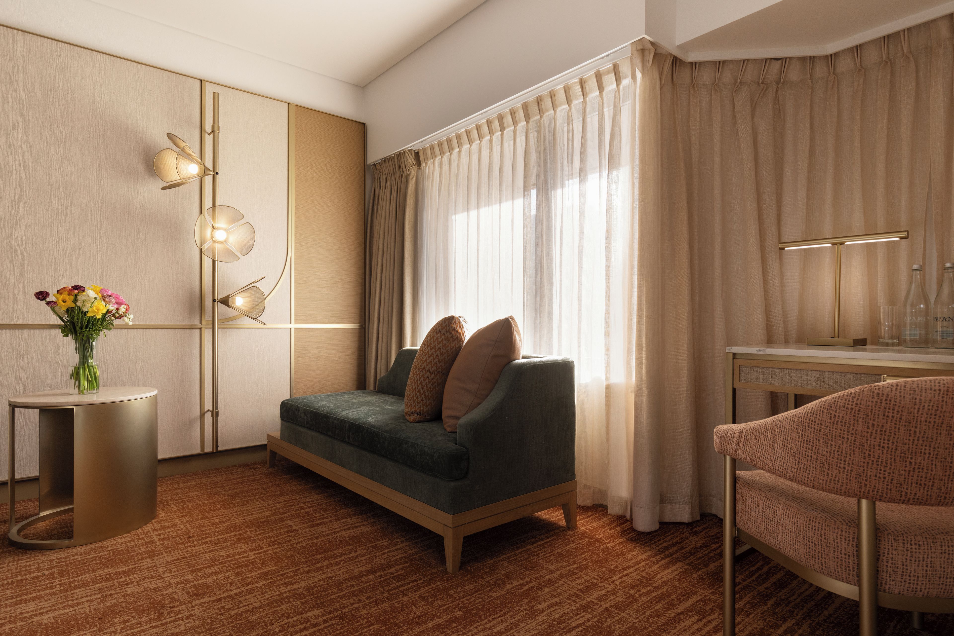 Quarto de hotel moderno com sofá, mesa lateral com um vaso de flores, escrivaninha com luminária e uma grande janela com cortinas.