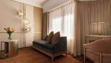 Quarto de hotel moderno com sofá, mesa lateral com um vaso de flores, escrivaninha com luminária e uma grande janela com cortinas.