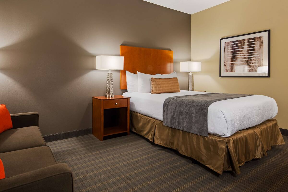 Quarto de hotel com cama, mesa de cabeceira, abajur, cadeira e um quadro na parede.